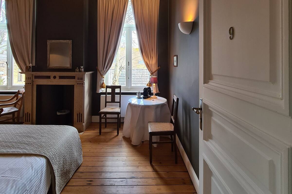 Quarto de hotel com piso de madeira, uma cama, uma mesinha com cadeiras, lareira e janela com cortinas.