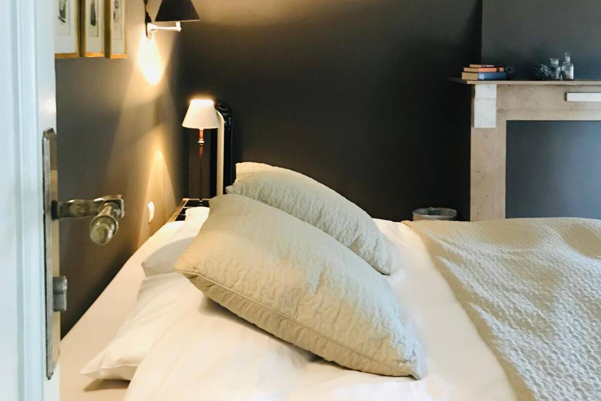 Um quarto com cama, luminária de cabeceira, lareira e quadros nas paredes.