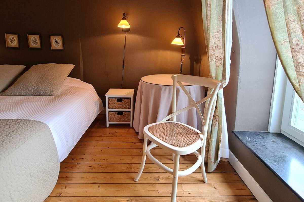 Quarto com piso de madeira, cama com roupa de cama branca, cadeira branca, pequena mesa com abajur e janela com cortinas.