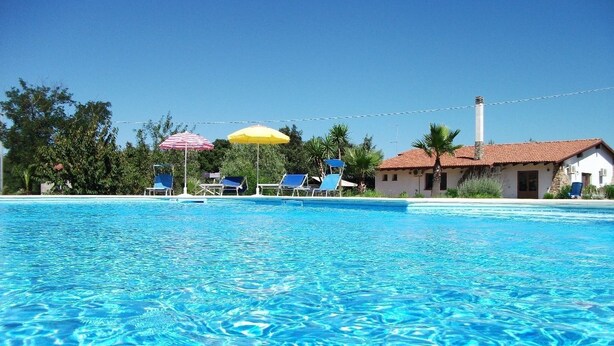 Uma piscina azul cristalina com espreguiçadeiras e guarda-sóis, um edifício branco com telhado vermelho e palmeiras ao fundo.