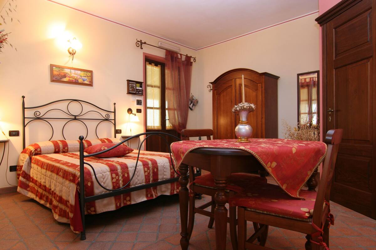 Um quarto com uma cama, uma mesa com cadeira, um guarda-roupa de madeira e um abajur fixado na parede.