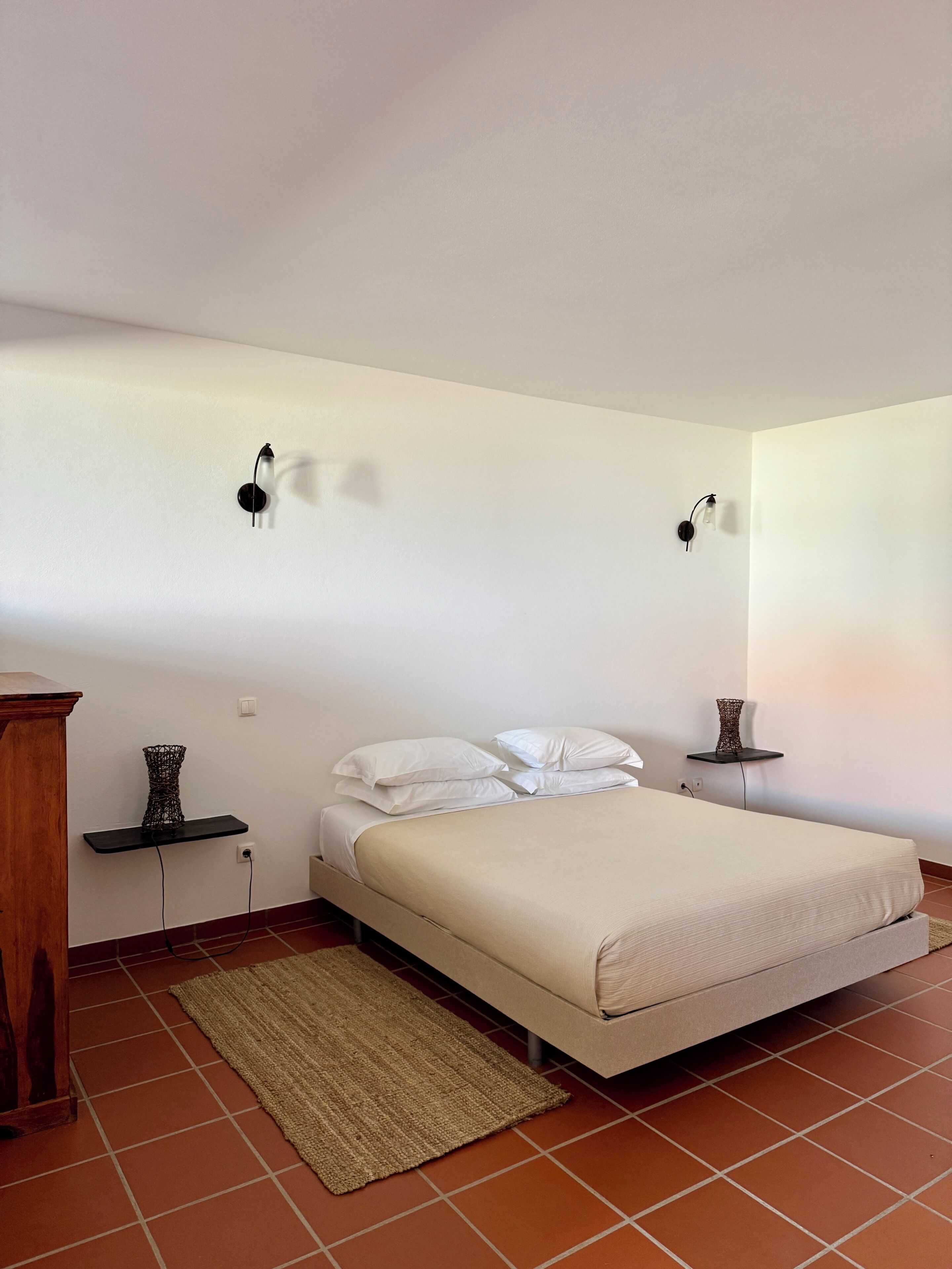 Quarto com cama, duas luminárias na parede, mesa de cabeceira, escrivaninha de madeira e tapete.