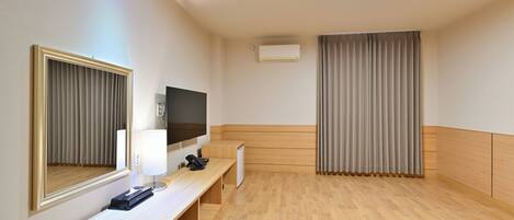 Quarto de hotel com piso de madeira, uma escrivaninha com telefone, um espelho, uma televisão e uma janela com cortinas.