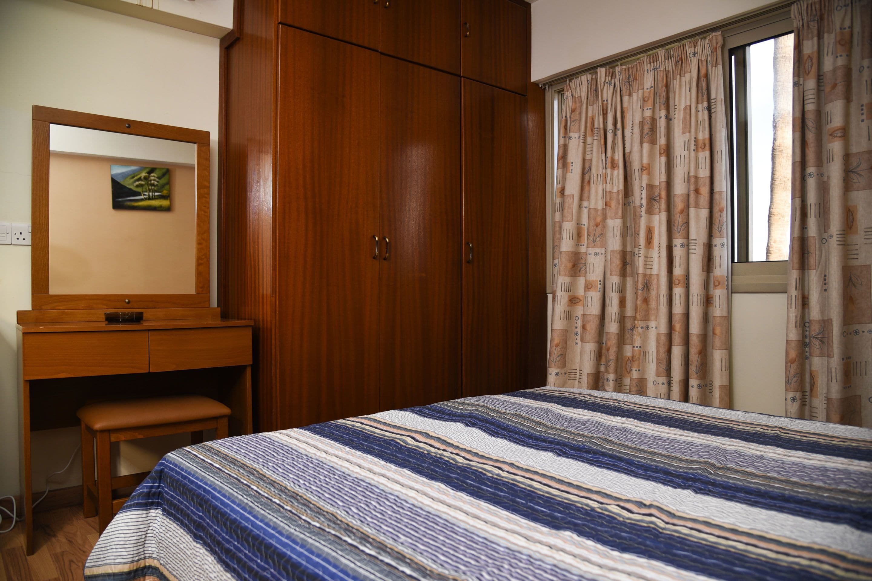 Quarto com cama, guarda-roupa de madeira, escrivaninha com cadeira e janela com cortinas.
