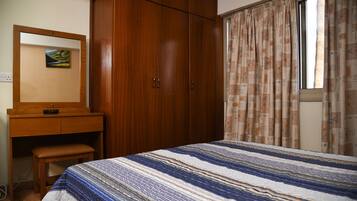 Quarto com cama, guarda-roupa de madeira, escrivaninha com cadeira e janela com cortinas.