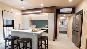 Uma cozinha moderna com ilha central, banquetas de madeira escura, micro-ondas embutido e geladeira de aço inoxidável.