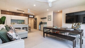 Uma sala de estar moderna com sofá, mesa de jantar com bancos, área de cozinha com geladeira e ventilador de teto.
