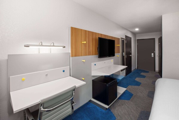 Quarto de hotel com cama, escrivaninha e cadeira, um refrigerador preto pequeno, televisão fixada em um painel de madeira e uma porta.