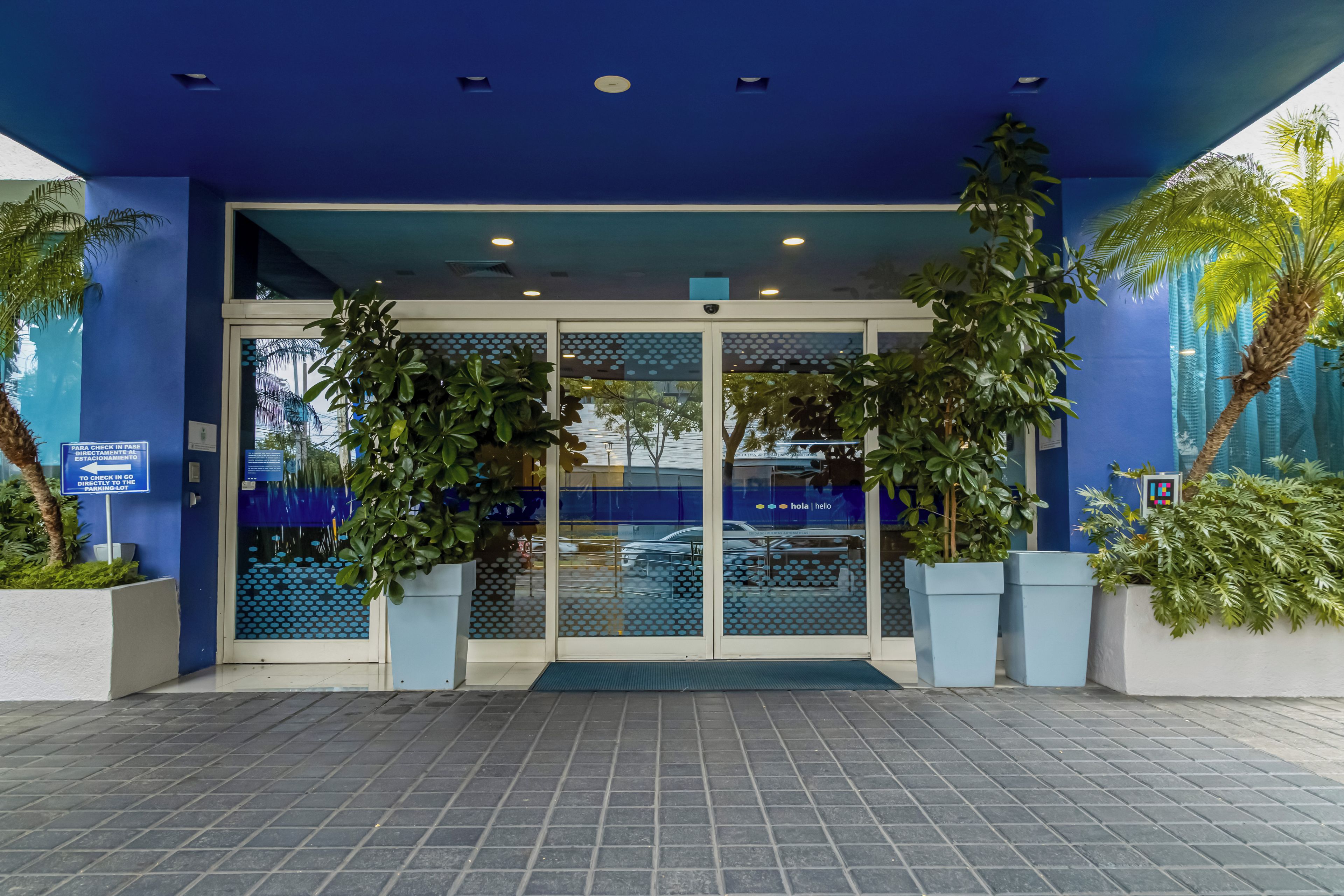 Entrada de hotel com fachada azul, portas de vidro e plantas em vasos.