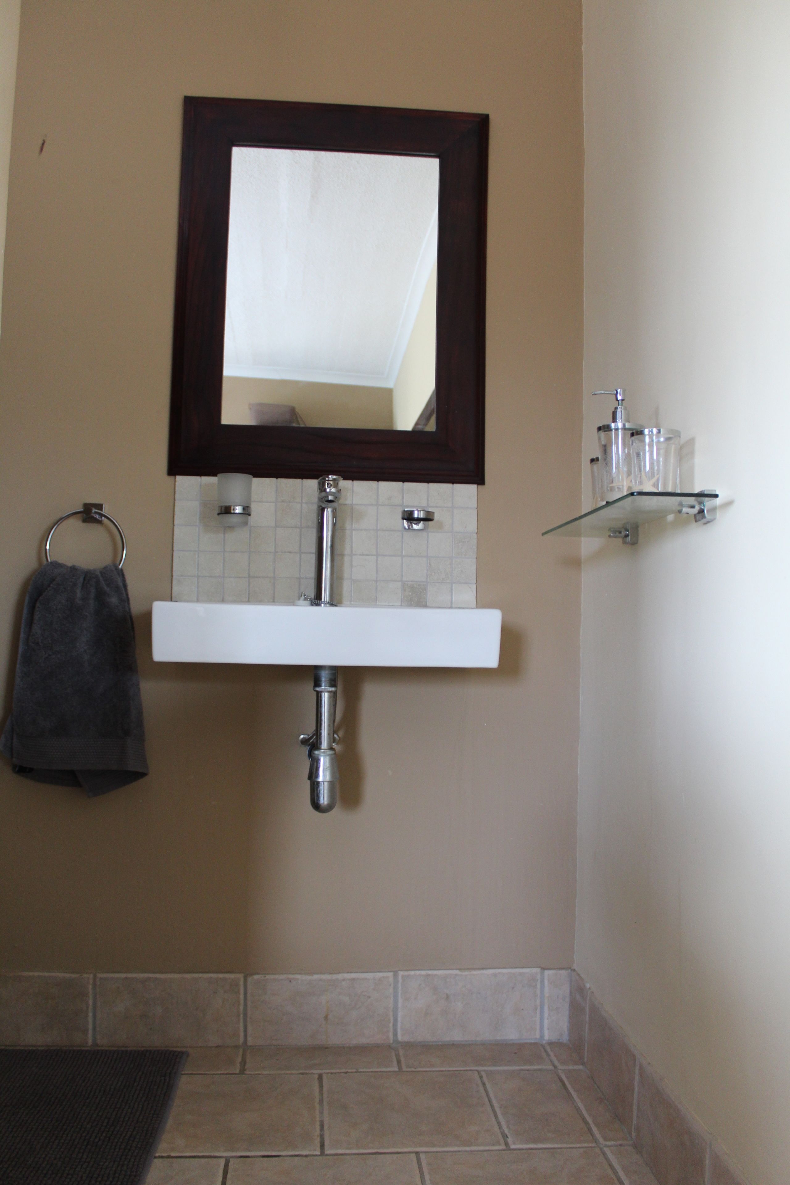 Casa de banho com lavatório branco, espelho, suporte para toalha e prateleira de vidro com artigos de higiene pessoal.