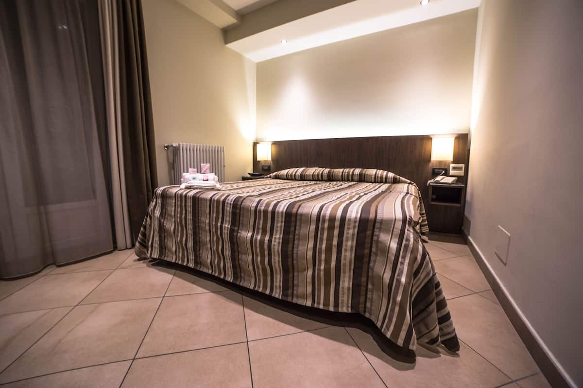 Quarto de hotel com uma cama grande, uma mesa de cabeceira, um abajur e um radiador.