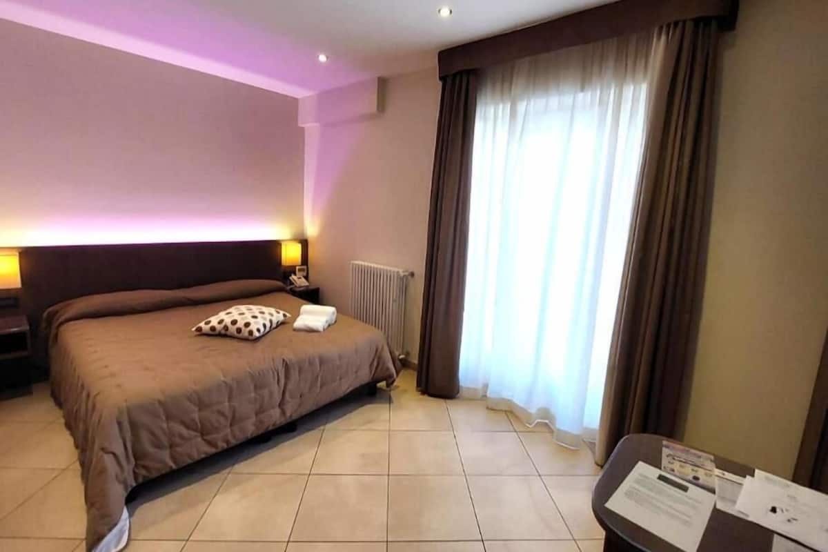 Quarto de hotel com uma cama grande, mesa de cabeceira com abajur, janela com cortinas e uma mesa com papéis.
