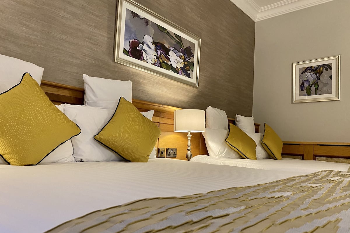 Quarto de hotel com uma cama grande, várias almofadas, uma mesa de cabeceira com abajur e dois quadros florais emoldurados na parede.