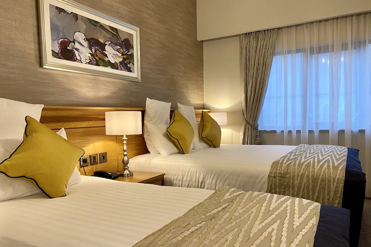 Quarto de hotel com duas camas, um quadro na parede, uma mesa de cabeceira com abajur e uma janela com cortinas.