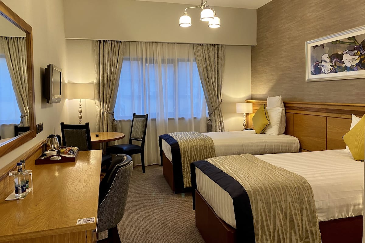 Quarto de hotel com duas camas, uma escrivaninha com cadeira, televisão e um quadro na parede.
