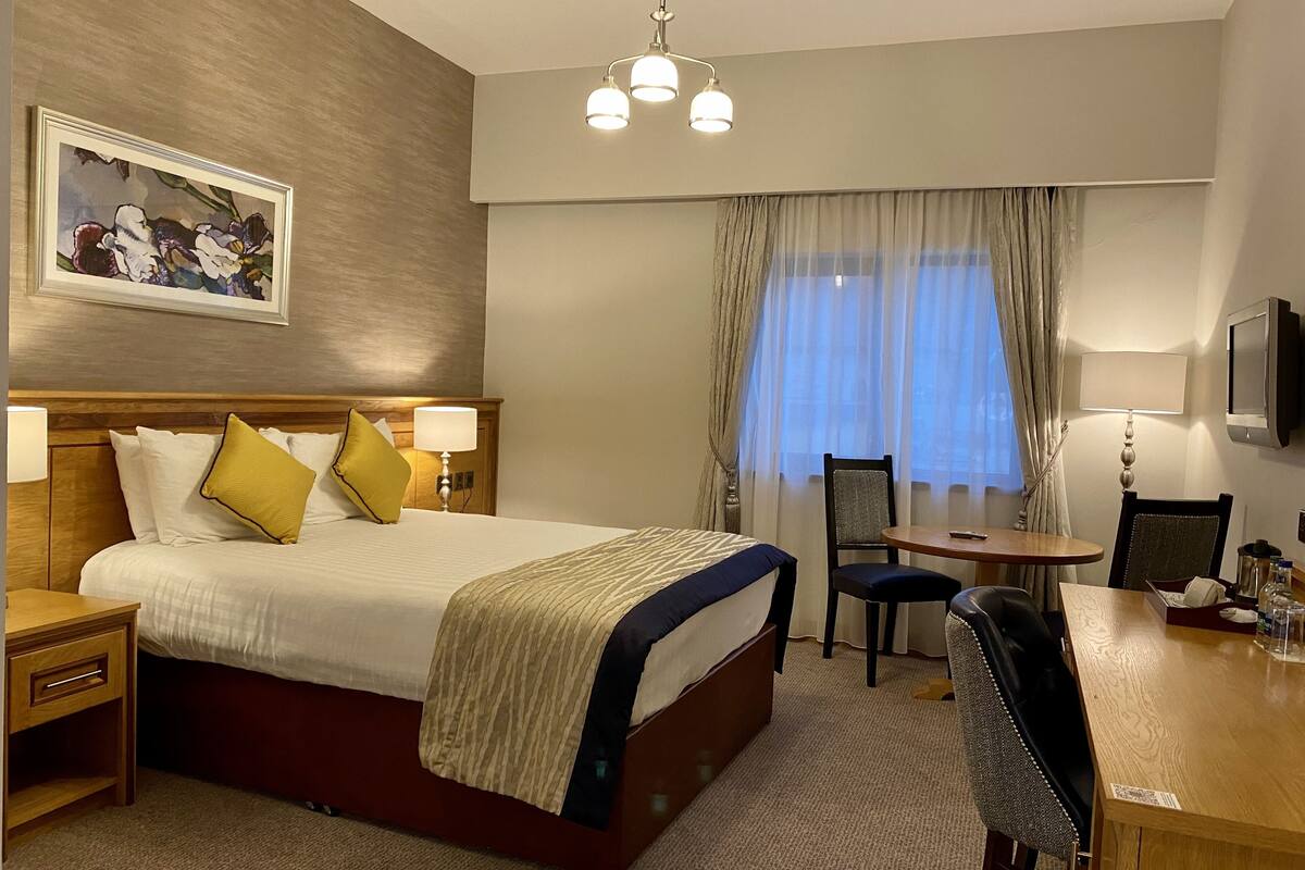 Quarto de hotel com cama, mesas de cabeceira, escrivaninha, cadeira, televisão e janela com cortinas.