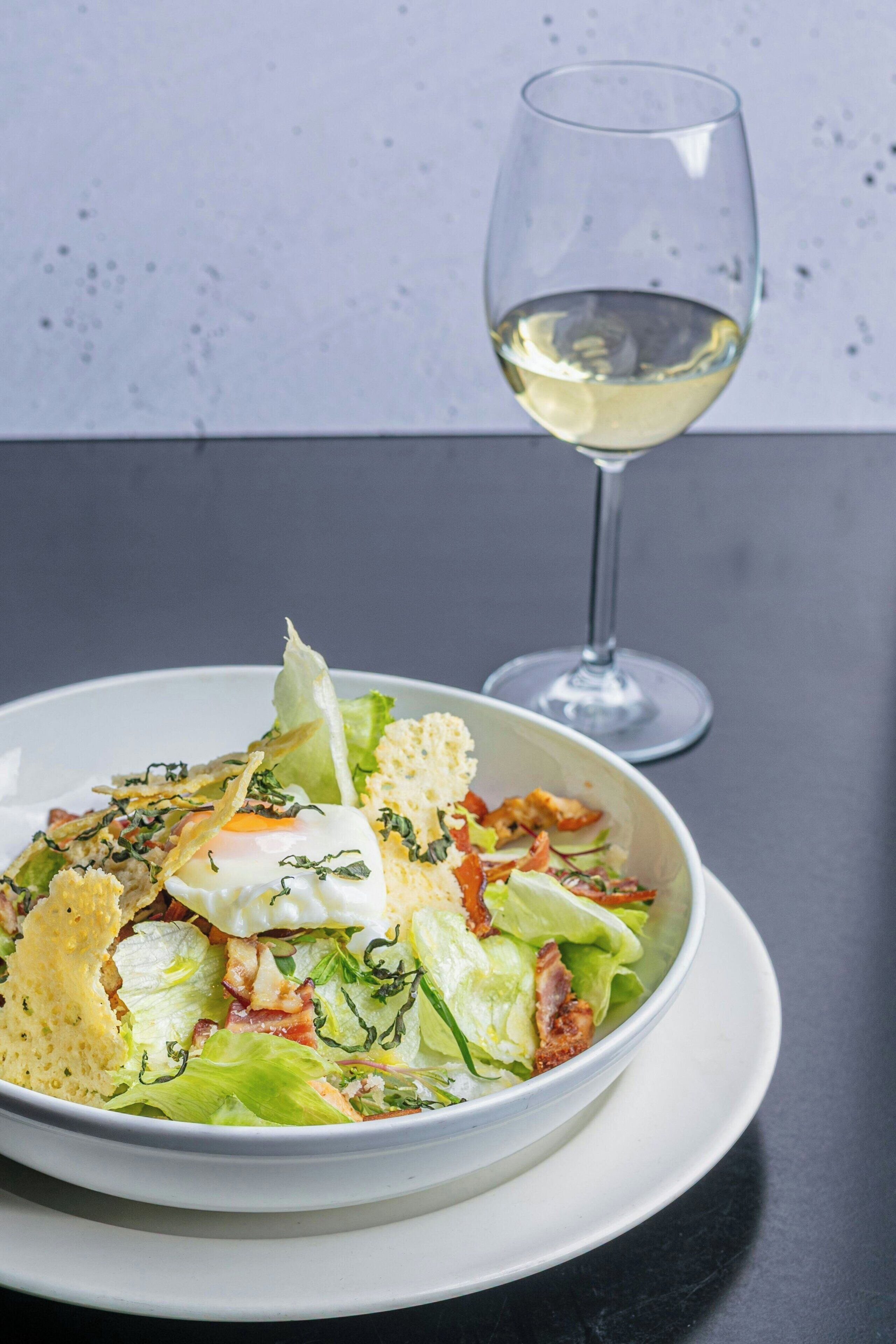 Uma salada com crôutons e ovo pochê numa travessa branca, acompanhada de um copo de vinho branco sobre uma mesa escura.