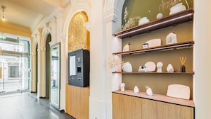 Uma área de recepção de hotel com balcão de check-in, terminal de autoatendimento e prateleiras decorativas com objetos como vasos e uma câmera.