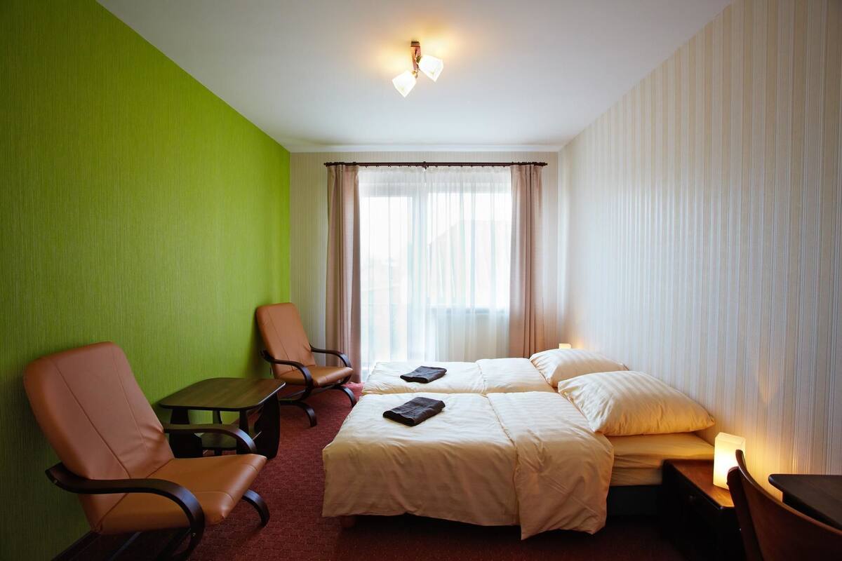 Quarto de hotel com duas camas, parede verde, janela com cortinas e uma mesinha.