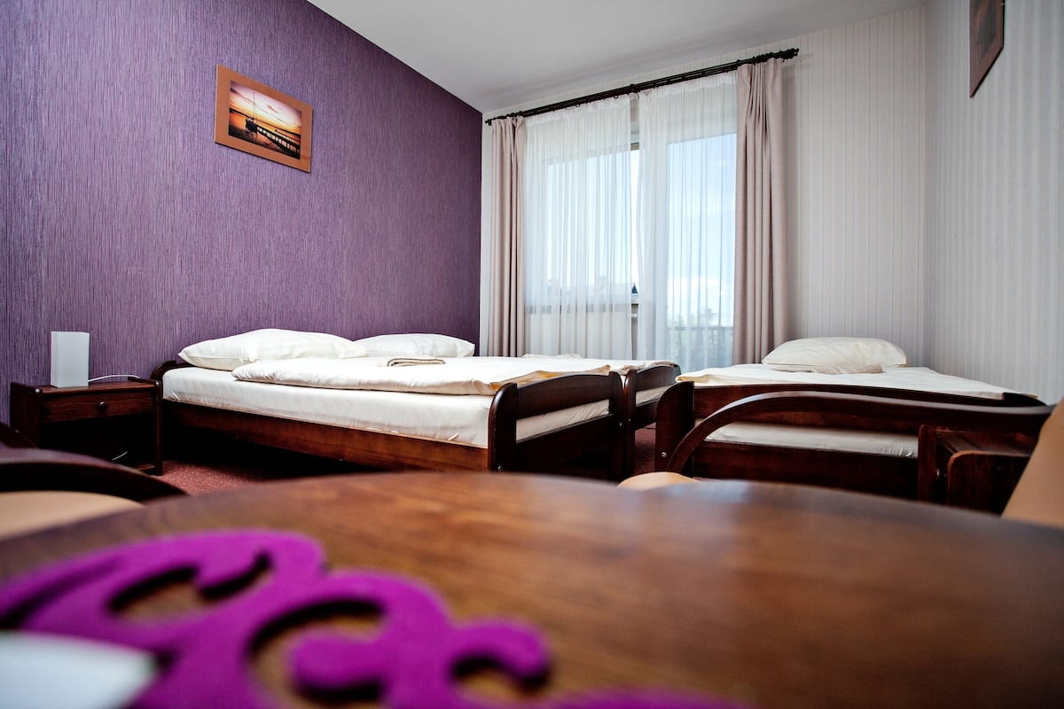 Quarto de hotel com duas camas de solteiro, uma mesa de madeira e parede roxa.