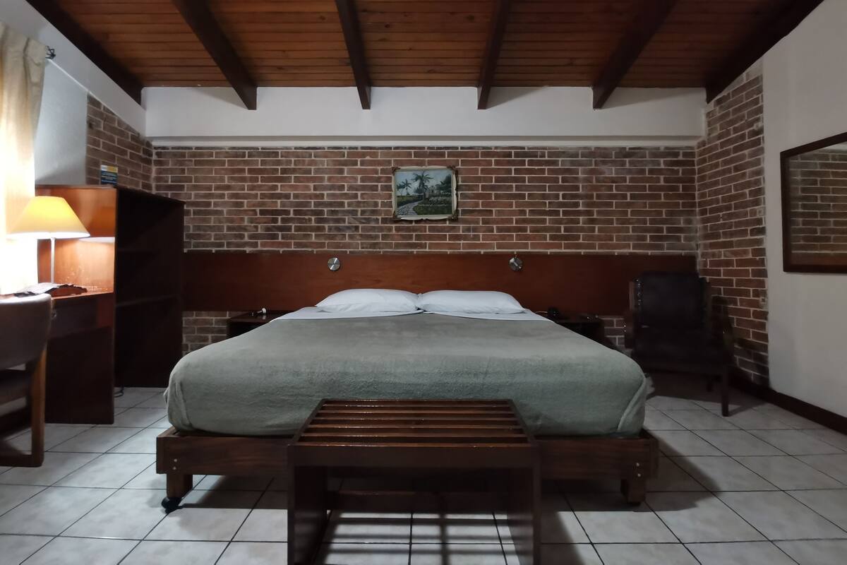 Quarto com cama de madeira, mesa de cabeceira, luminária e parede de tijolos.
