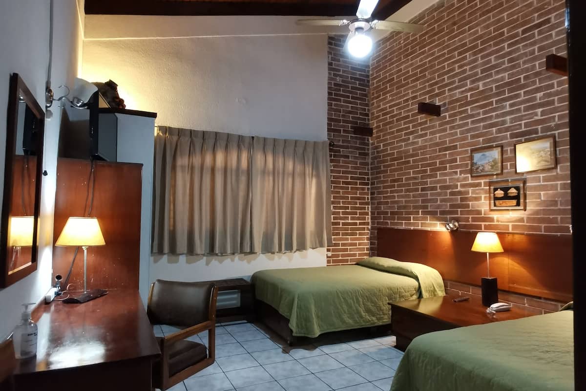 Quarto de hotel com parede de tijolos, duas camas, uma escrivaninha com cadeira, um espelho e um ventilador de teto.