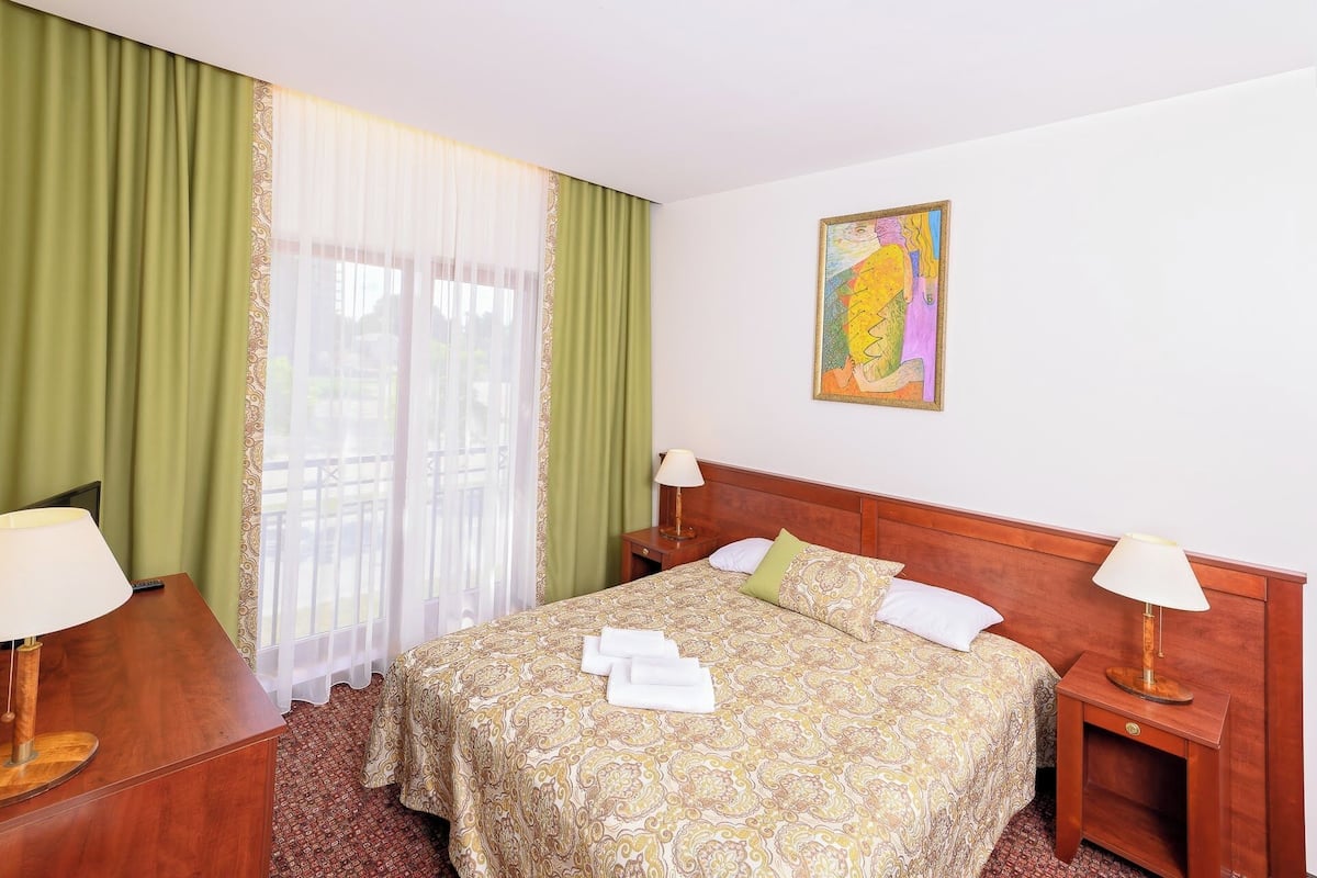 Quarto de hotel com uma cama grande, duas mesas de cabeceira com luminárias, um quadro na parede e uma janela com cortinas leves.