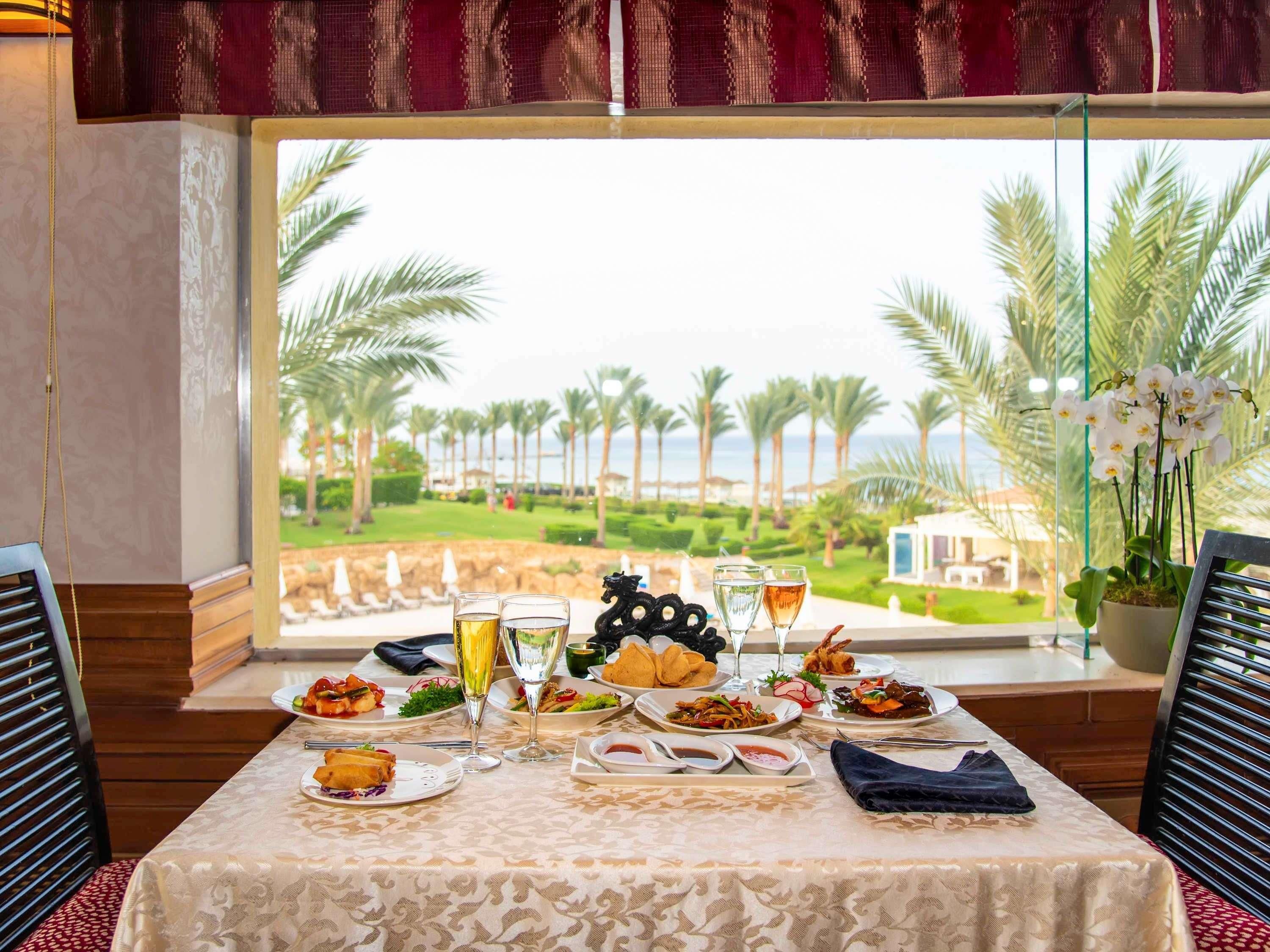 Uma mesa de jantar com diversos pratos e bebidas, diante de uma janela com vista para palmeiras e uma praia.