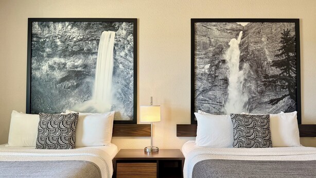 Quarto de hotel com duas camas, cada uma com almofada estampada, mesa de cabeceira com abajur e duas grandes pinturas em preto e branco de cachoeiras na parede.