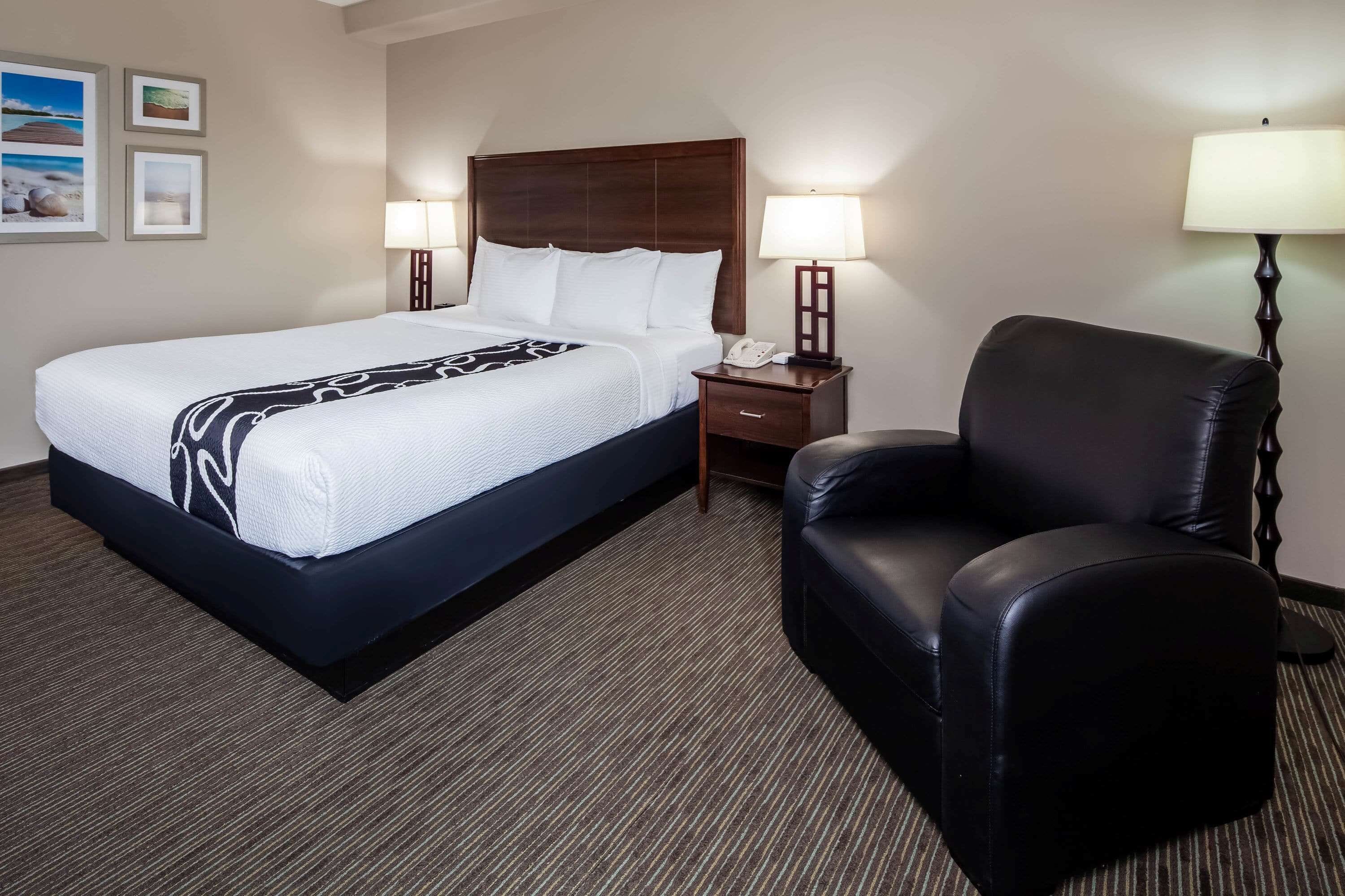 Quarto de hotel com uma cama grande, duas mesas de cabeceira com luminárias, uma poltrona de couro preto e quadros nas paredes.