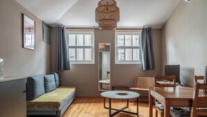 Quarto com sofá, mesa redonda, mesa de jantar com cadeiras, espelho e duas janelas com cortinas.