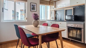 Uma área de refeições com mesa de madeira e cadeiras vermelhas, uma cozinha com micro-ondas e forno, e uma vista pela janela.
