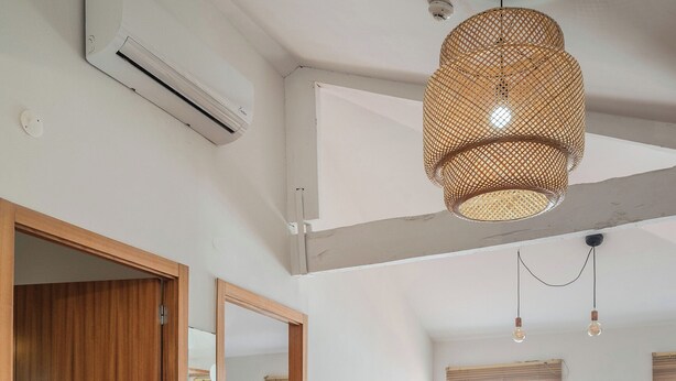 Um quarto espaçoso com piso de madeira, área de refeições com mesa e cadeiras, luminária pendente de tear e um aparelho de ar condicionado na parede.