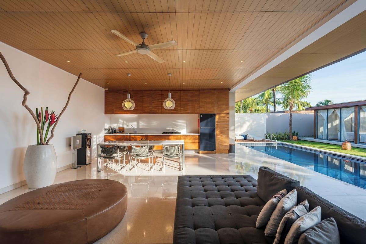 Uma sala de estar moderna com um sofá grande, uma mesa de centro e uma área de jantar com cadeiras e uma mesa. Visível ao fundo, há uma piscina.