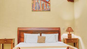 Quarto com cama de madeira, um banquinho, mesa de cabeceira, abajur, pintura e lustre.
