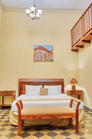 Quarto com cama de madeira, um banquinho, mesa de cabeceira, abajur, pintura e lustre.