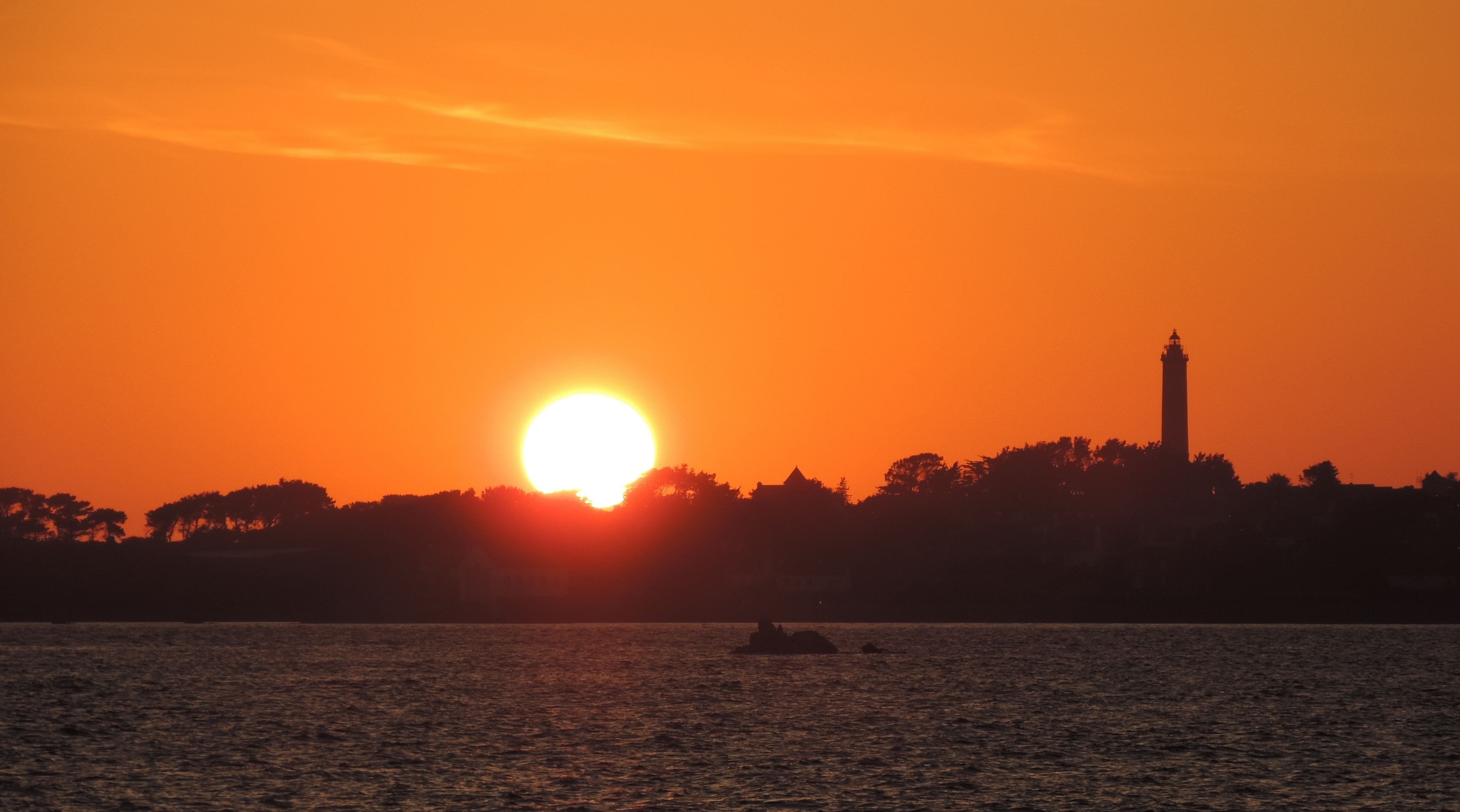Pôr do sol sobre a água, com silhuetas de uma torre e árvores.