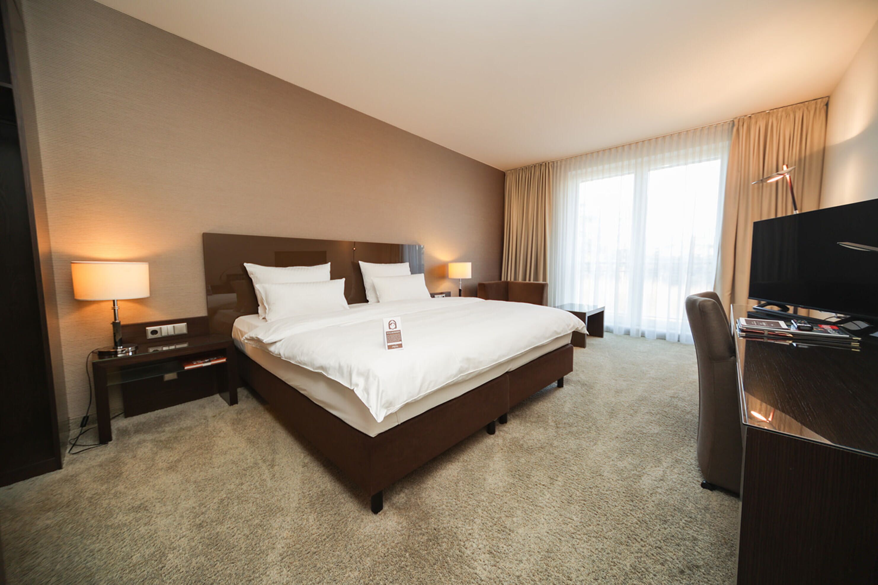 Quarto de hotel com uma cama grande, duas mesas de cabeceira com abajures, uma escrivaninha com uma TV e vista para a cidade pela janela.