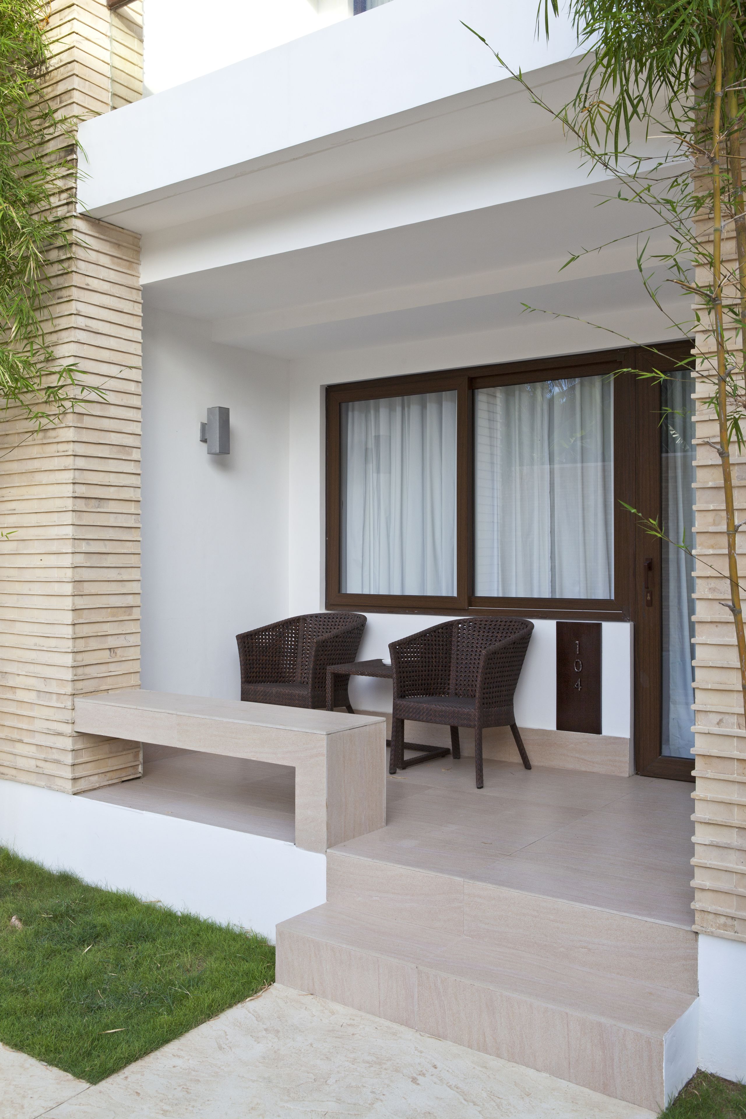 Uma área de estar moderna ao ar livre, com duas cadeiras de vime e um banco, uma parede externa branca e uma janela grande com cortinas.