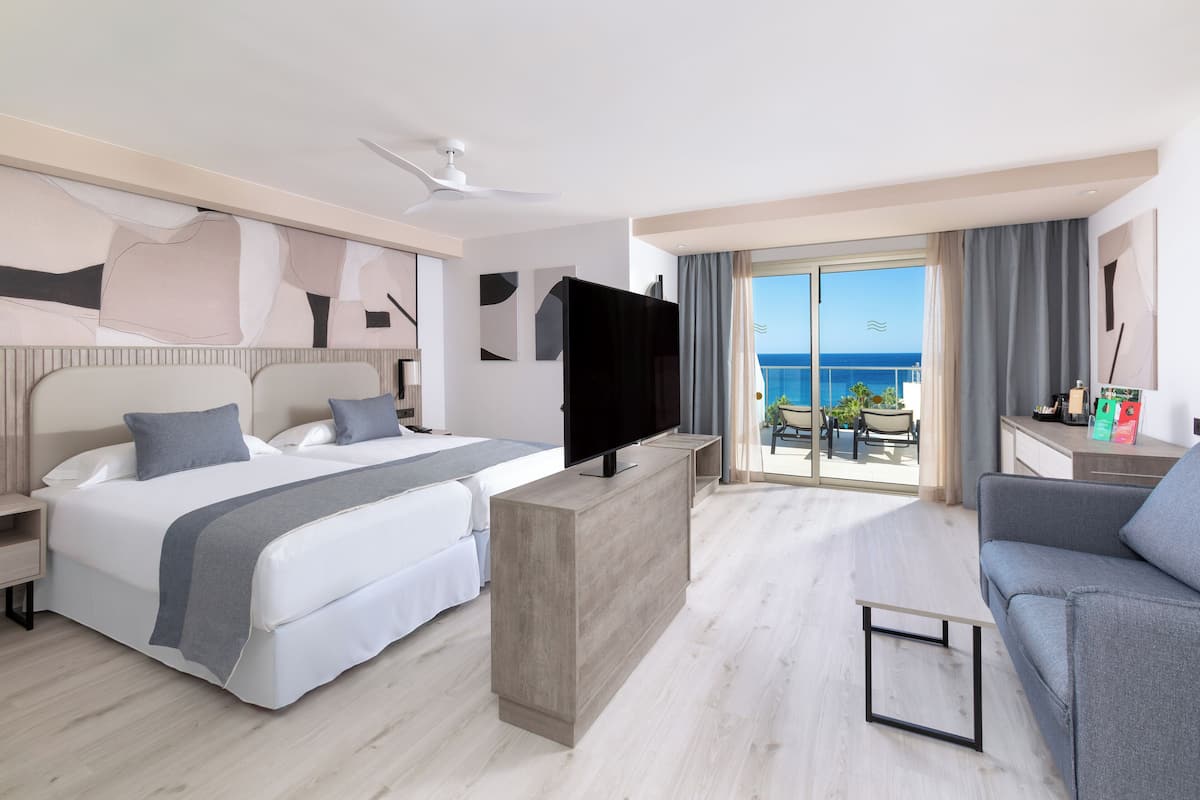 Quarto de hotel moderno com cama, televisão, sofá e varanda com vista para o mar.