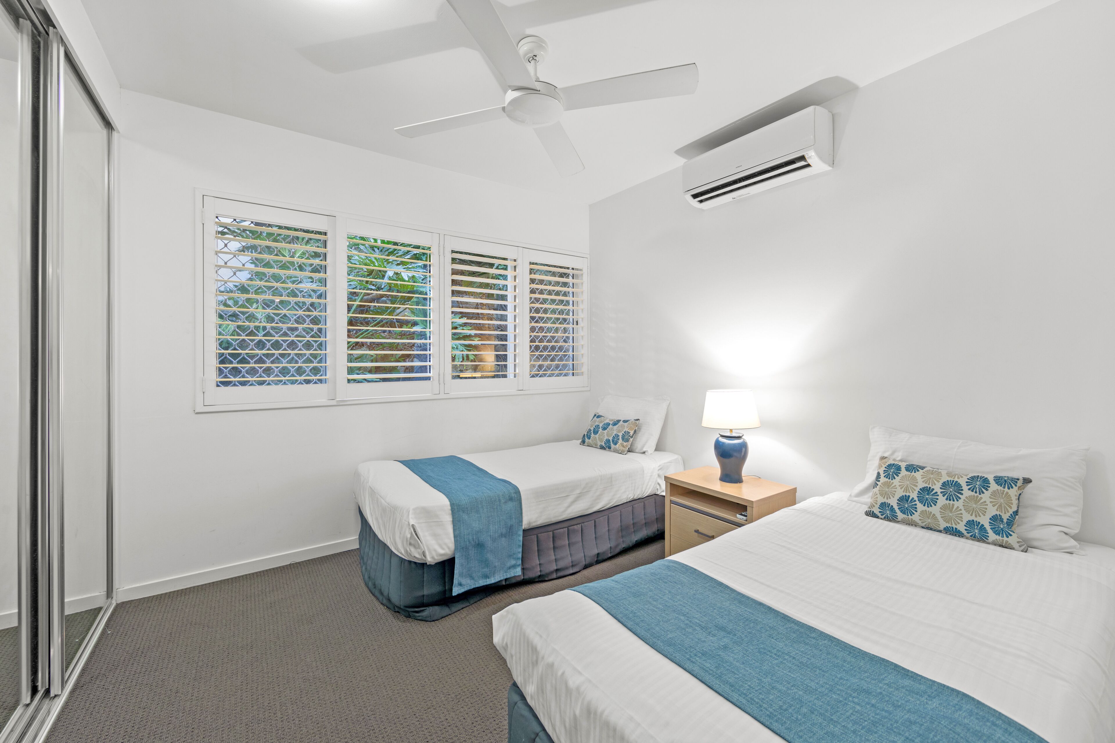 Quarto com duas camas, ventilador de teto, ar condicionado e janela com persianas.