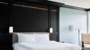 Quarto de hotel com uma cama grande, duas luminárias de cabeceira, uma cadeira e vista para a cidade.