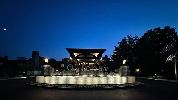 Uma entrada bem iluminada com design arquitetônico moderno, destacando um pavilhão central e área ajardinada com espelho d'água.