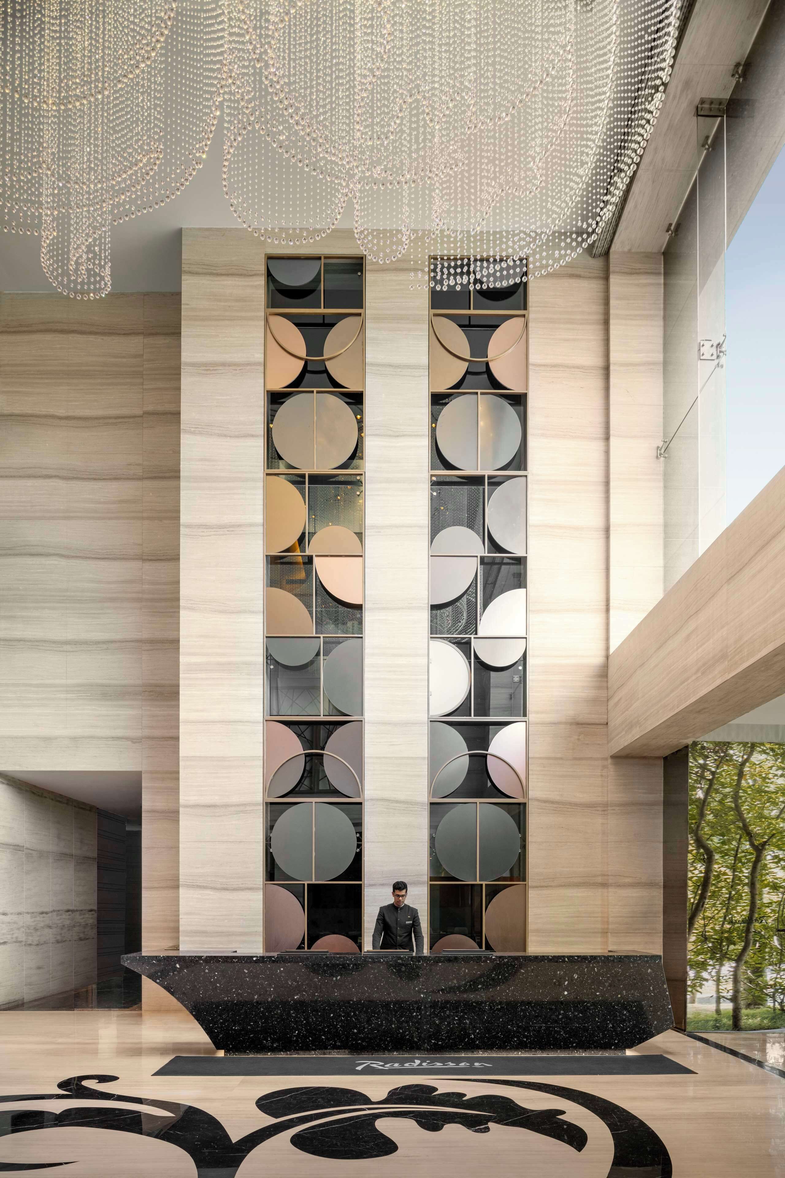 Uma área de recepção moderna, com uma grande parede com padrão geométrico, um balcão de recepção preto e um candelabro.