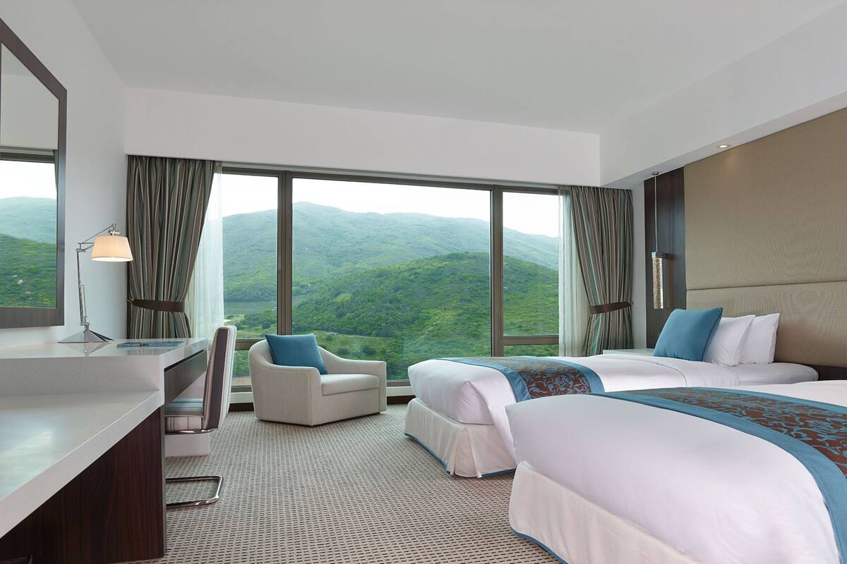 Quarto de hotel com duas camas, uma escrivaninha, uma cadeira e janelas amplas com vista para as montanhas.