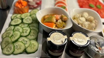 Mesa de buffet com diversos alimentos, incluindo pepinos fatiados, uma tigela de legumes e dois potes de comida conservada.