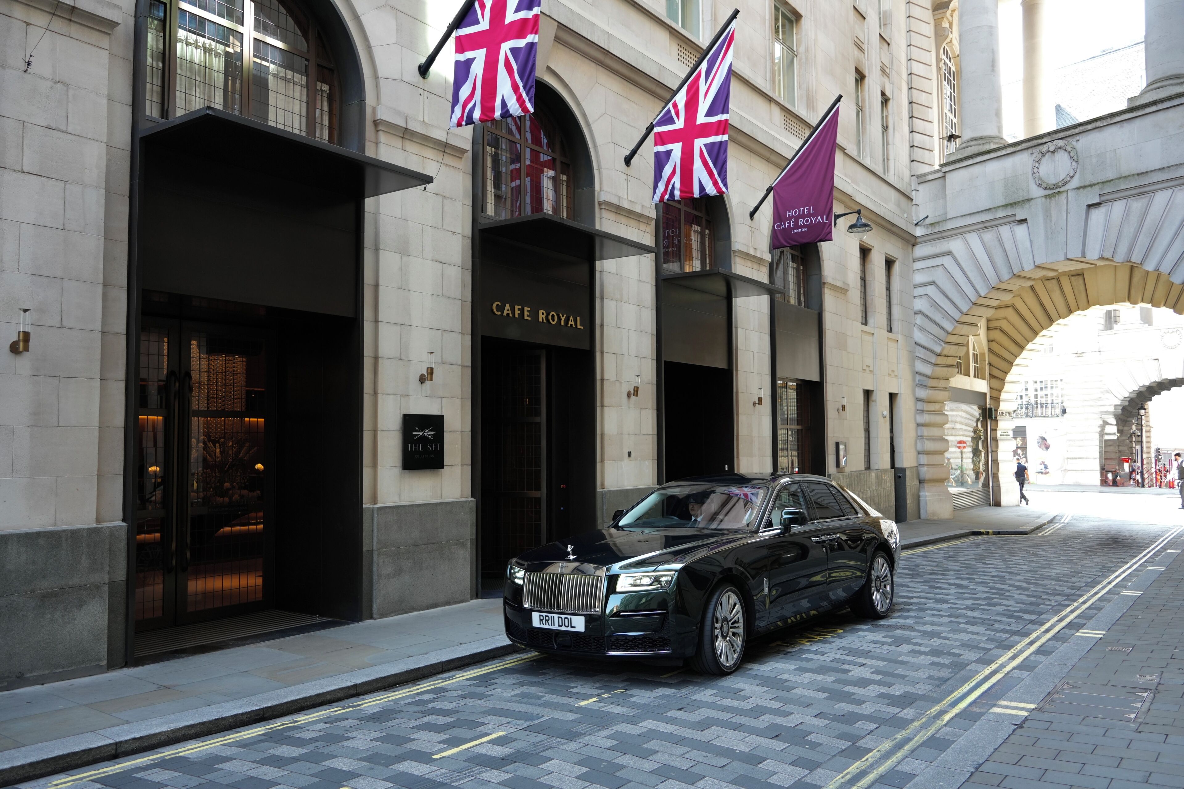 Um carro de luxo preto estacionado em frente a um edifício com bandeiras e uma placa que diz 'CAFE ROYAL'.