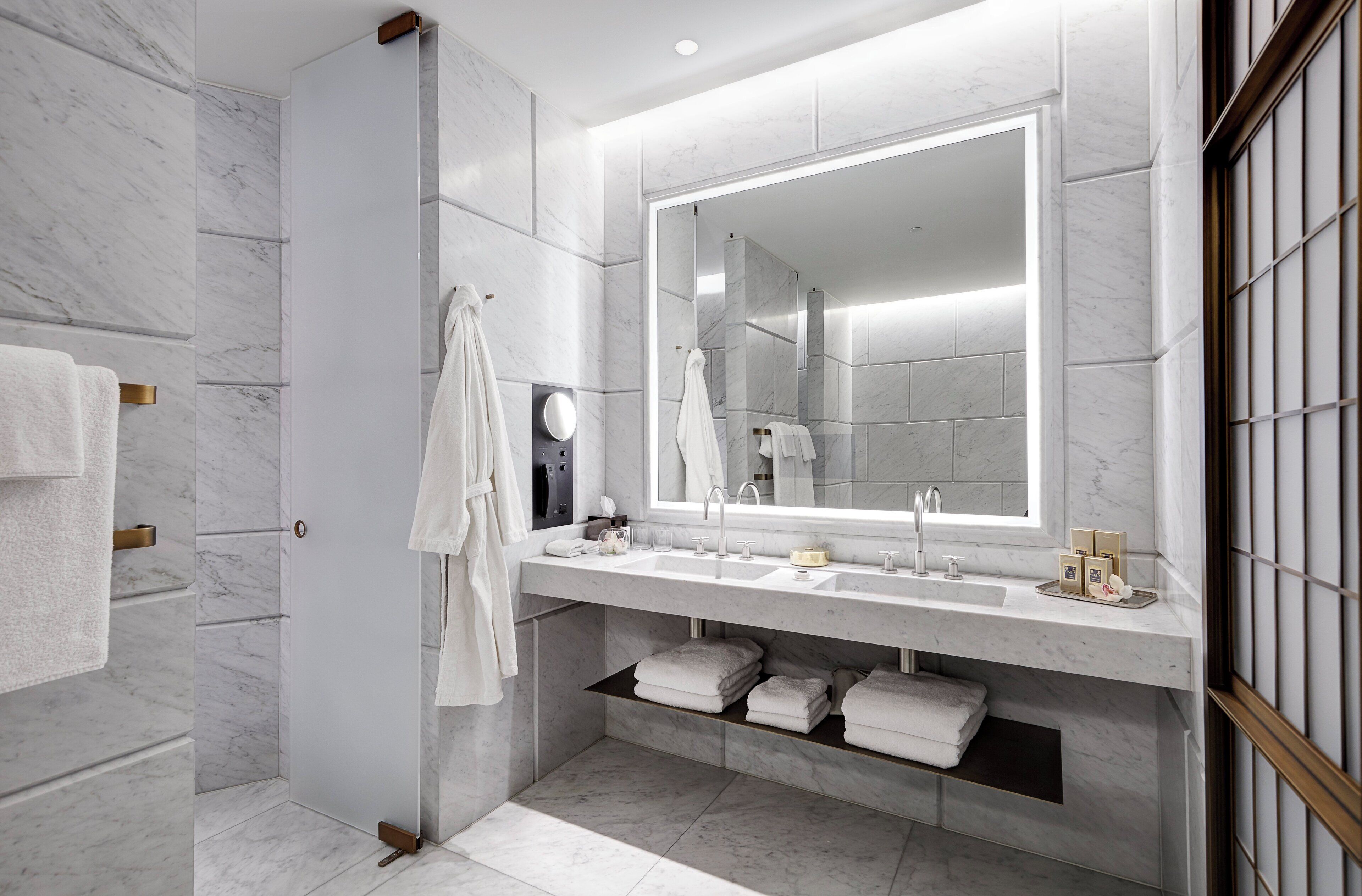 Um banheiro moderno com um espelho grande, azulejos de mármore branco, chuveiro embutido e toucador com pia e espaço para toalhas.