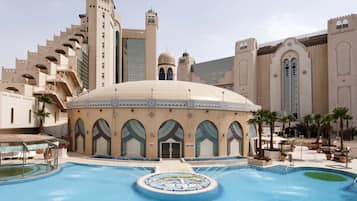 Uma piscina grande com uma fonte central, rodeada por palmeiras e um edifício com janelas em arco e uma cúpula.