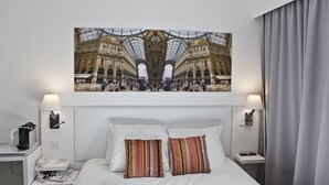 Quarto de hotel com uma cama grande, duas almofadas, uma mesa de cabeceira com abajur, telefone e um quadro com uma cena movimentada de rua na parede.